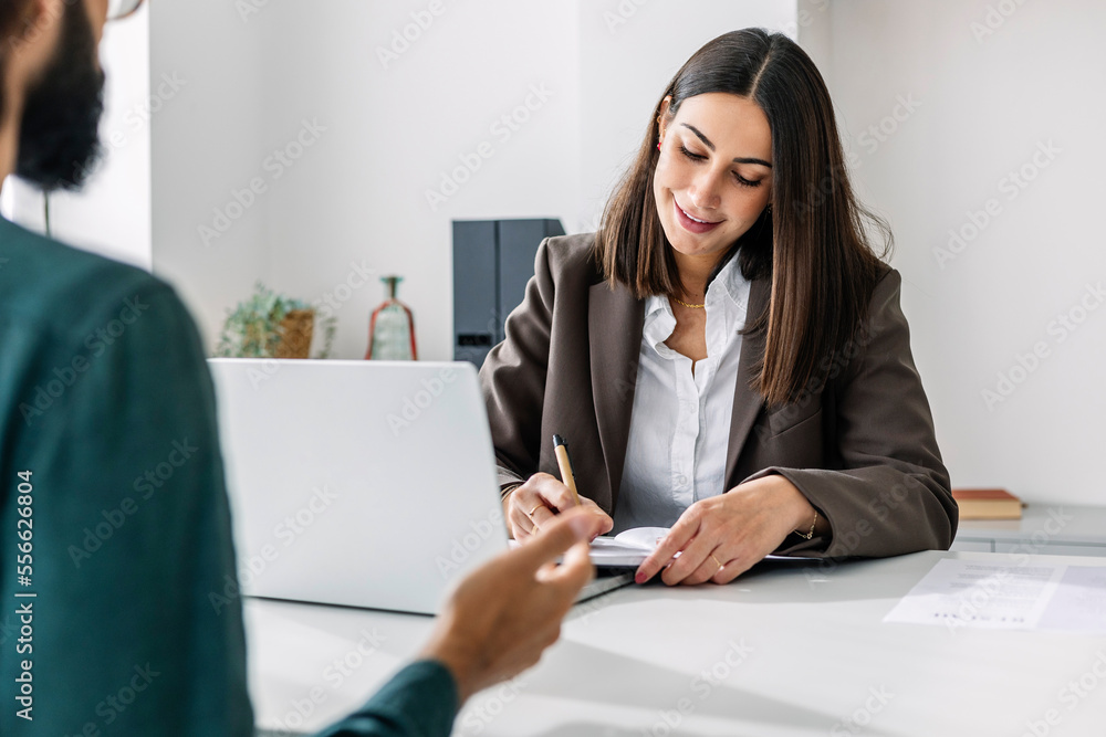 Smiling recruiter interviewing candidate and writing in diary at desk ...
