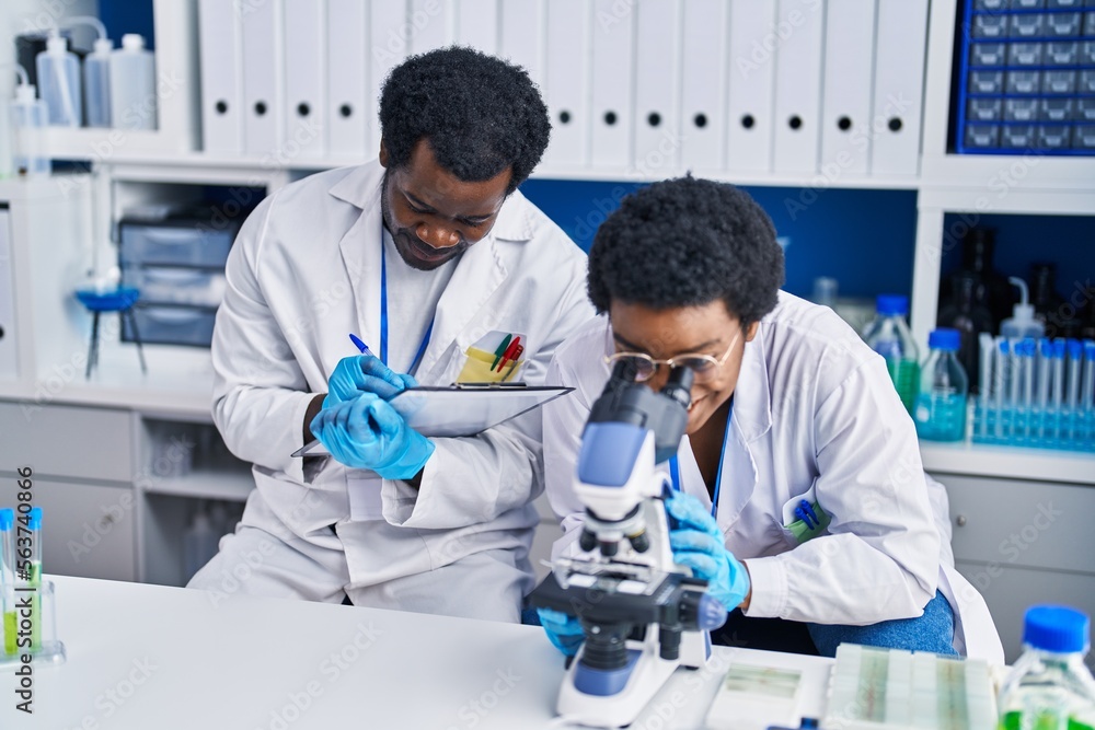 African american man and woman scientists using microscope writing on document at laboratory ...