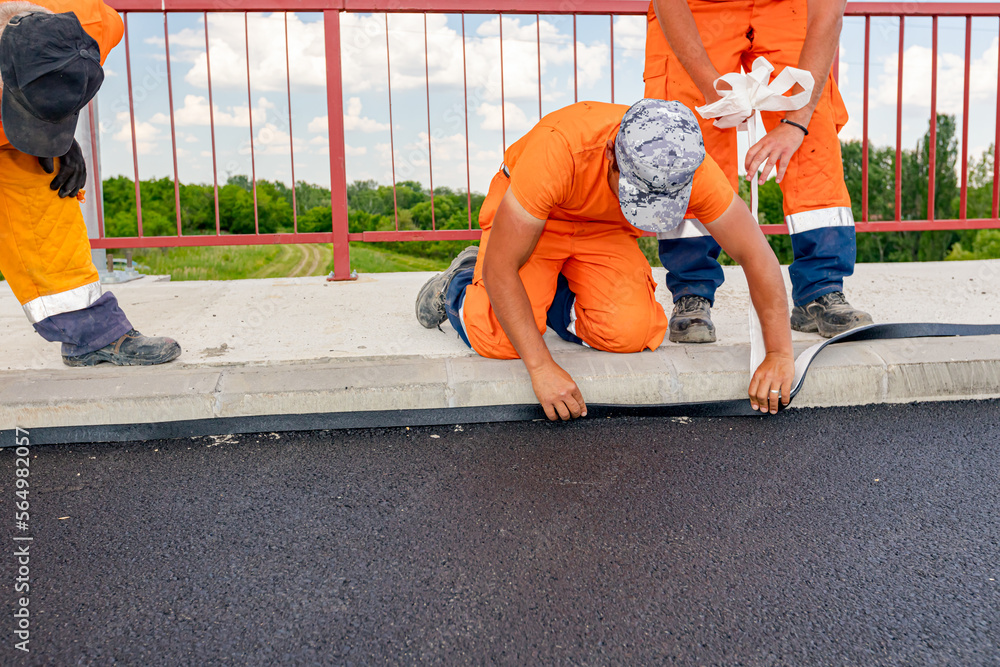 Workers apply black bitumen membrane strips for waterproofing roads and ...