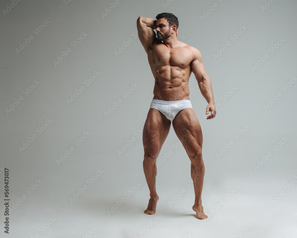 Sexy muscular man in white underwear posing in studio. Handsome bodybuilder standing on grey ...