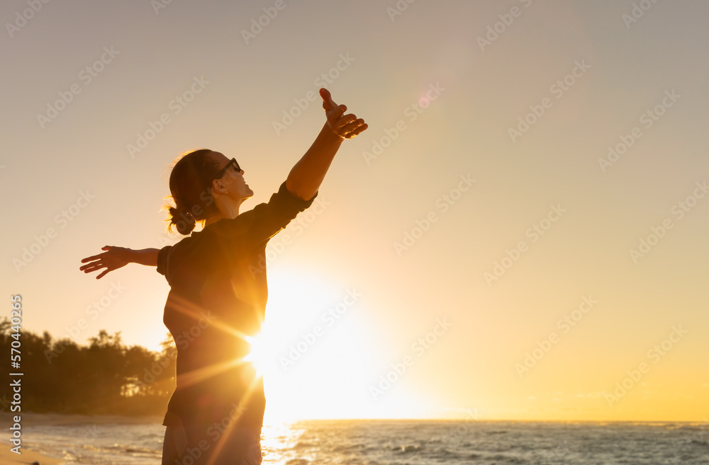 Young woman facing ocean sunset rejoices, laughs, smiles looking up to ...
