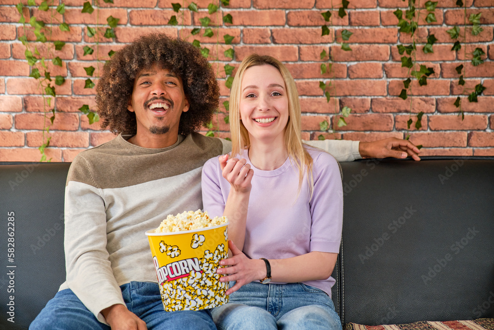 young multiracial couple sitting on the sofa at home watching tv and ...