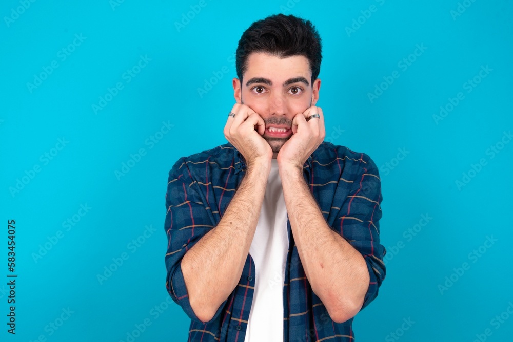 Fearful Young man standing over blue studio background keeps hands near ...