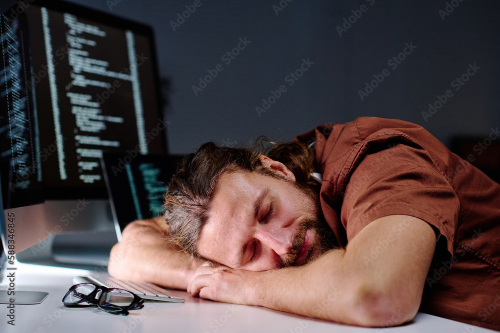 Young exhausted male programmer with his head on desk by computer keyboard sleeping after work ...