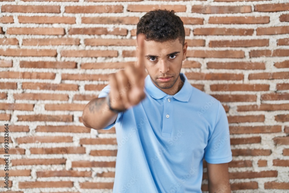 Brazilian young man standing over brick wall pointing with finger up ...