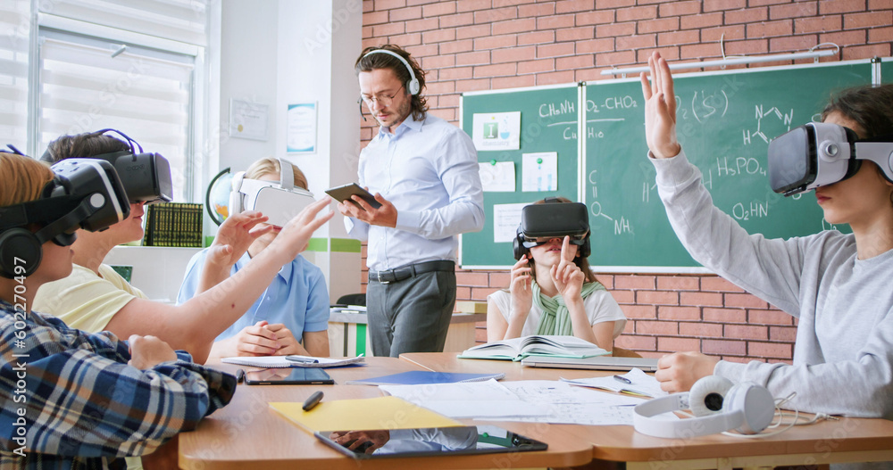 Group of young pupils in VR headsets sitting together at desk while ...