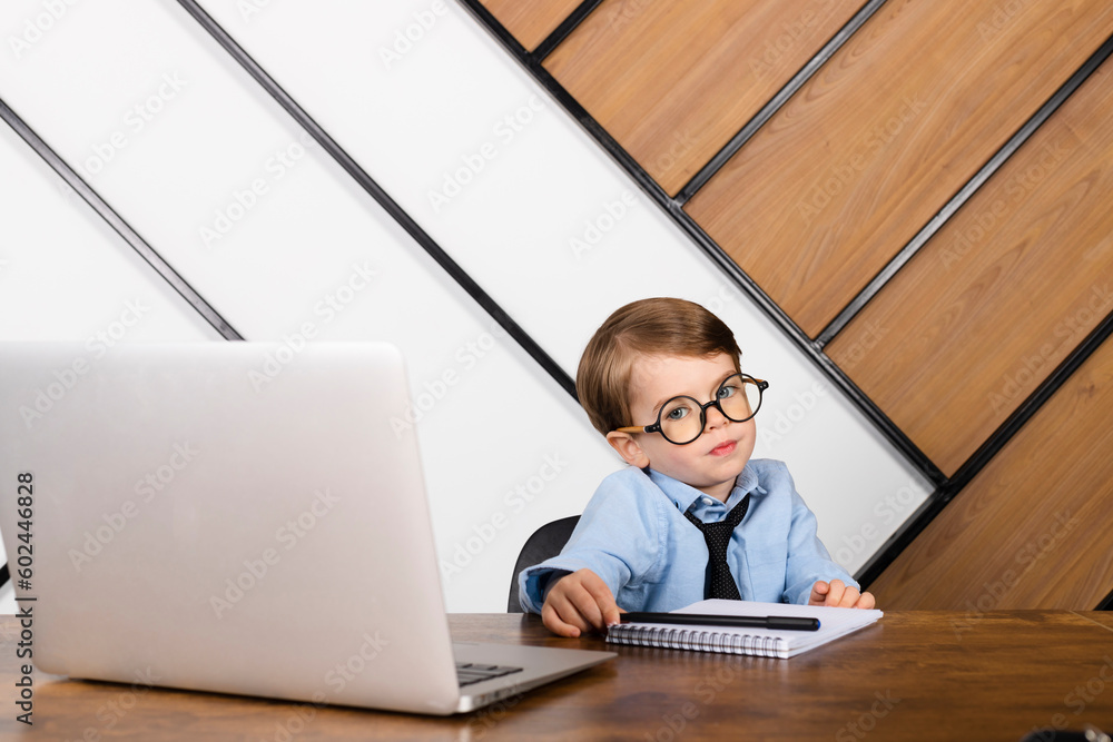 Little boy in round eyeglasses, blue shirt and tie sitting at the desk ...