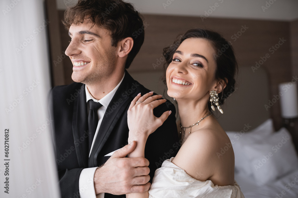 joyful bride in elegant jewelry and wedding dress hugging shoulder of ...