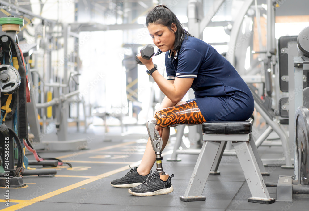 Woman with prosthetic leg sitting in gym lifting dumbbell weight. Asian ...