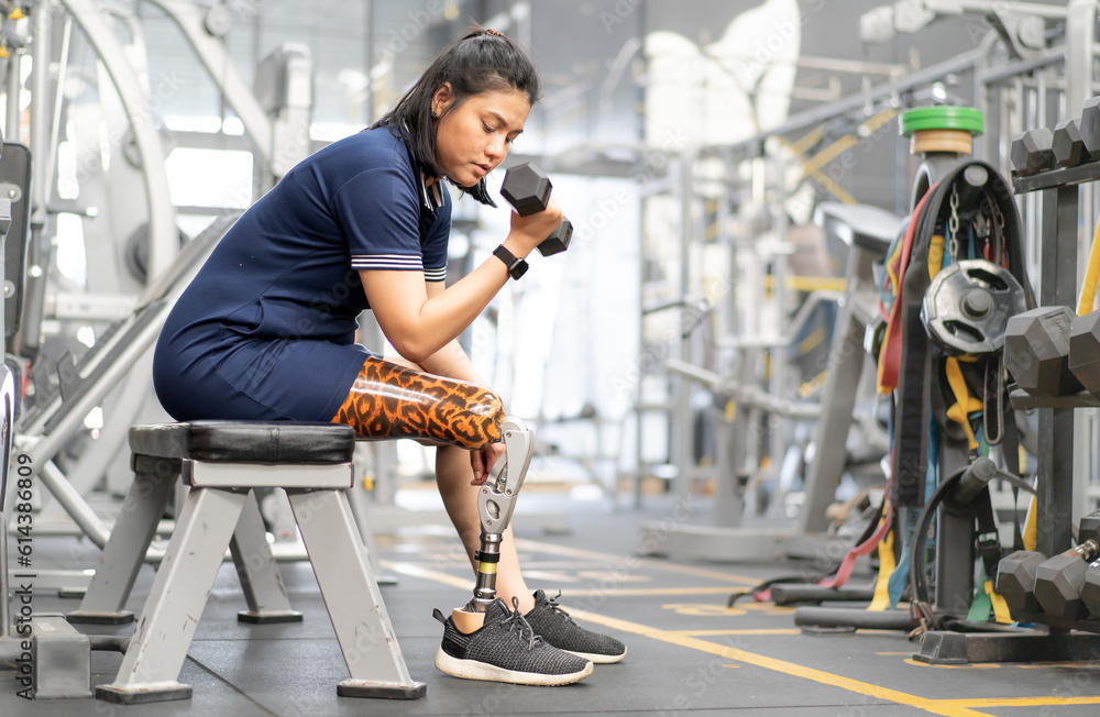 Woman with prosthetic leg sitting in gym lifting dumbbell weight. Asian ...