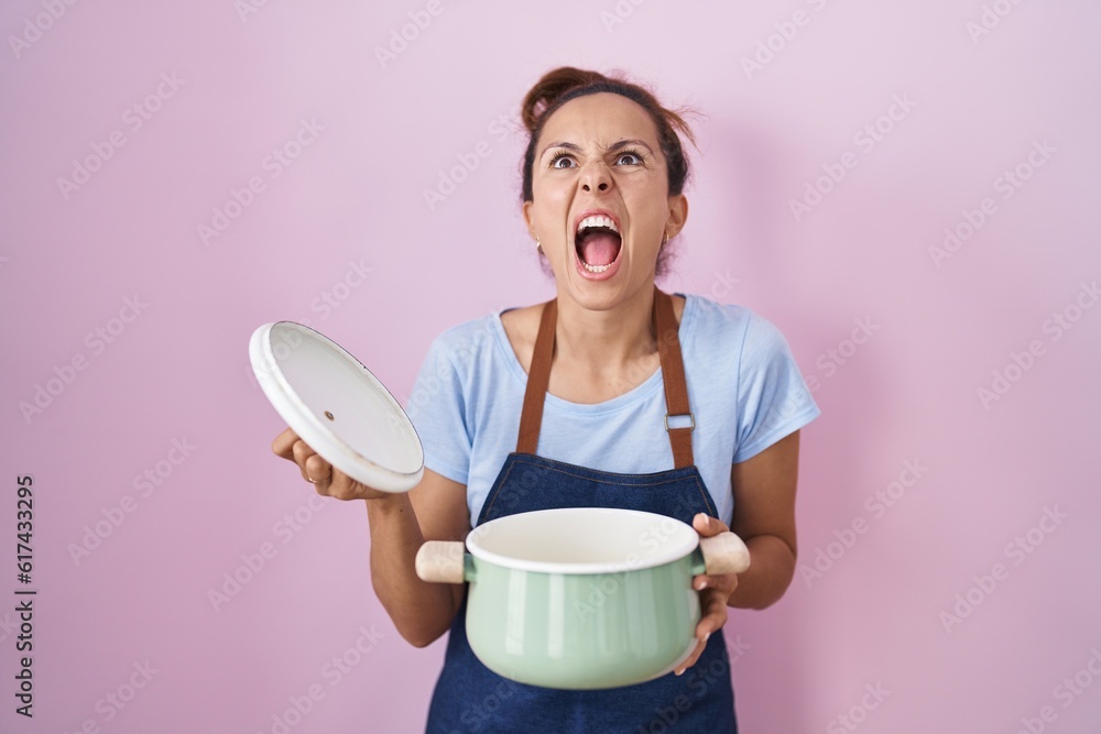 Brunette woman wearing apron holding cooking pot angry and mad ...