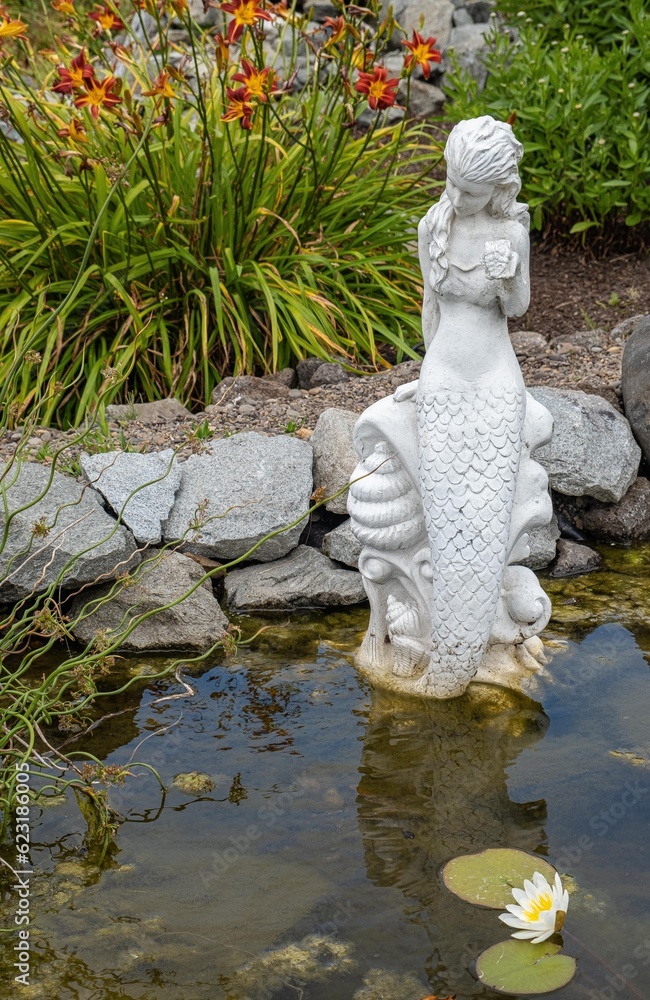 A tranquil stone statue sculpture of a mermaid overlooks a still pond ...