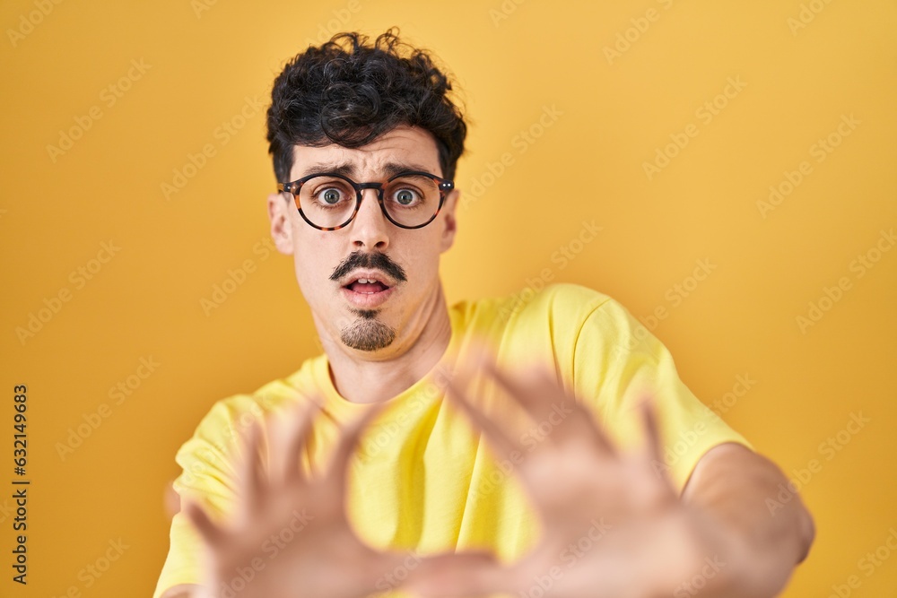 Hispanic man wearing glasses standing over yellow background afraid and ...