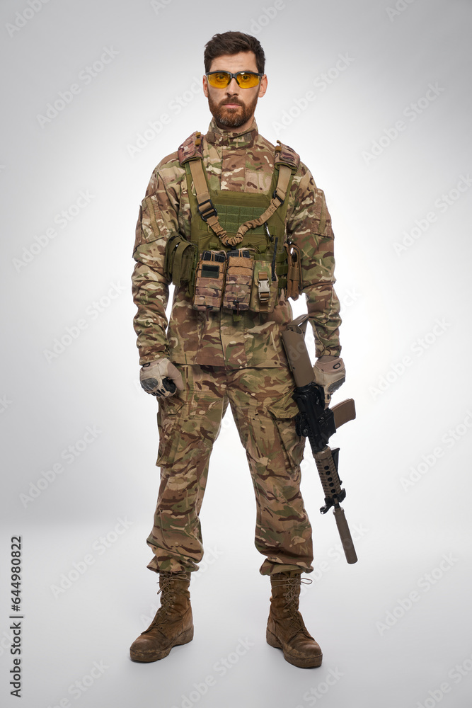 Serious male soldier in body armor holding rifle, while posing indoors ...
