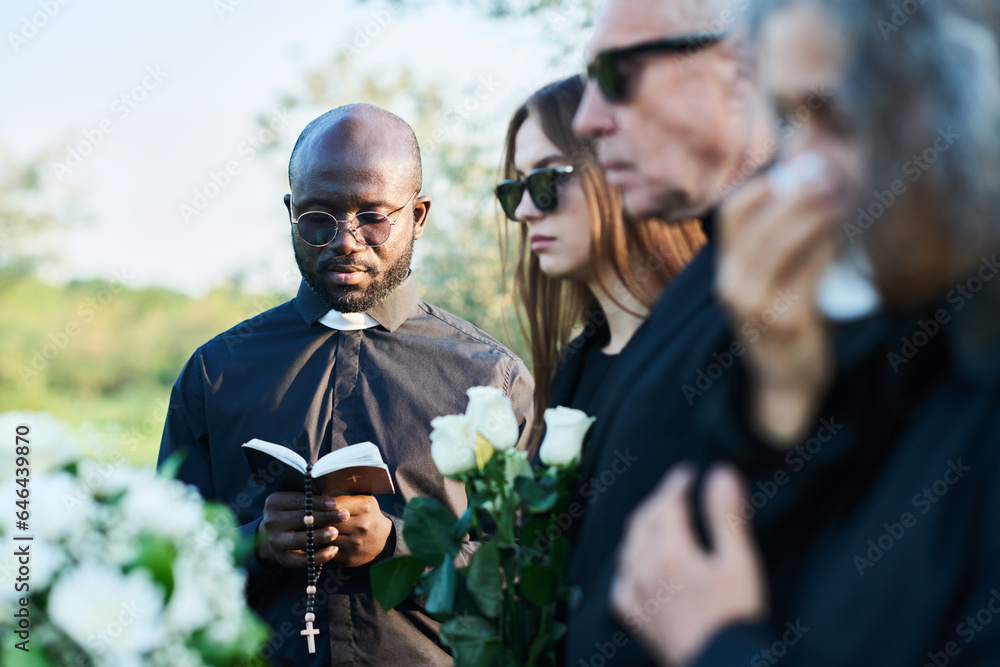 Focus on serious priest in black attire looking in open Holy Bible at ...