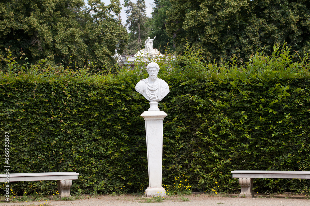 A tall trimmed hedge, in front of it stone benches and a bust on a ...