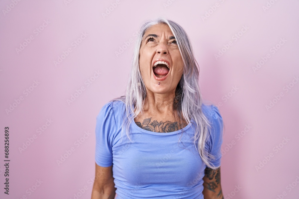 Middle age woman with tattoos standing over pink background angry and ...