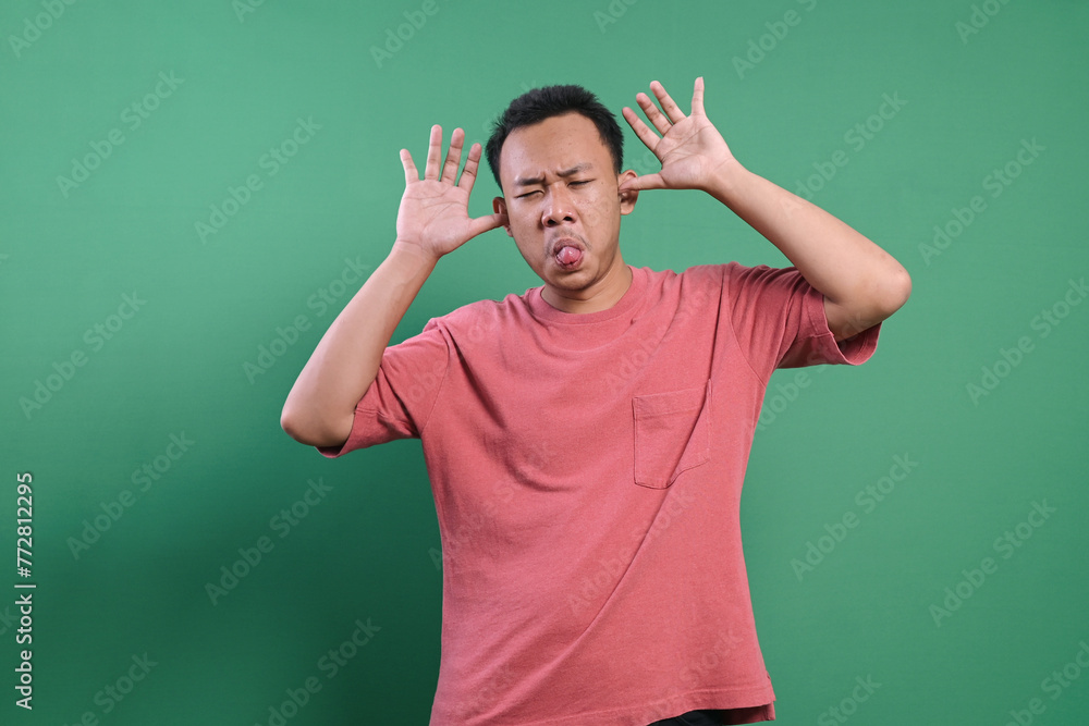 Portrait of silly Asian male making funny faces with hands on face and ...