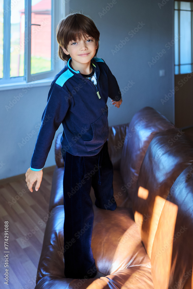 7 year old European boy stands on leather sofa in full height in his ...