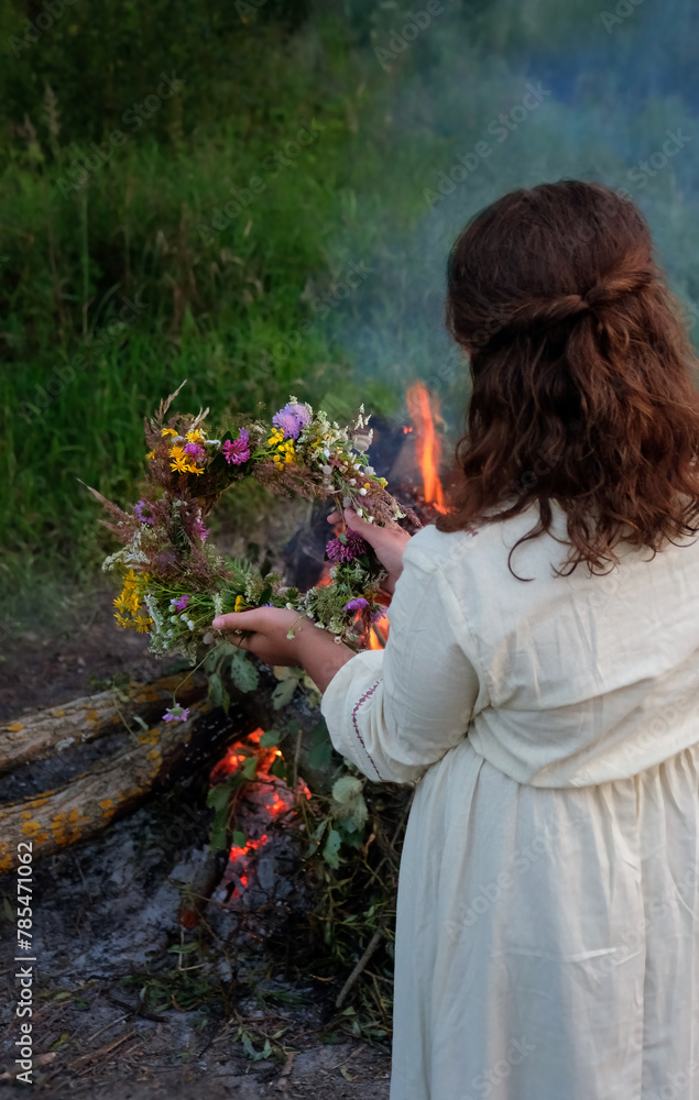 Girl with floral wreath in hands near campfire outdoor, dark natural background. rear view ...