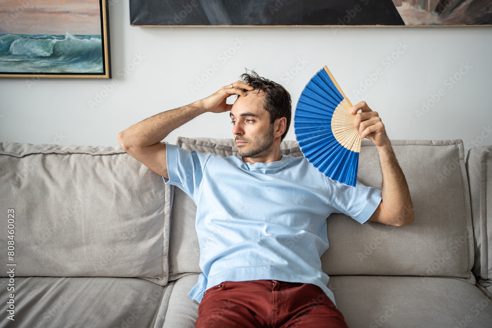 Tired man with fan suffering from heat. Guy sprawled on couch with ...