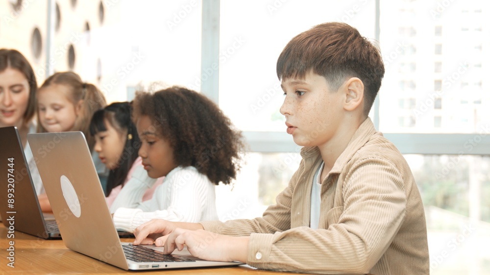 Boy playing laptop with multicultural friend learning prompt at STEM technology class ...