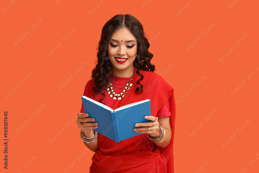 Beautiful young Indian woman in sari reading book on orange background ...