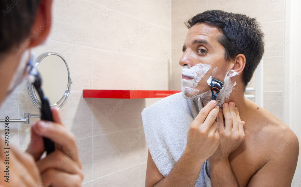 Young white man shaving with his razor in the bathroom at home looking ...