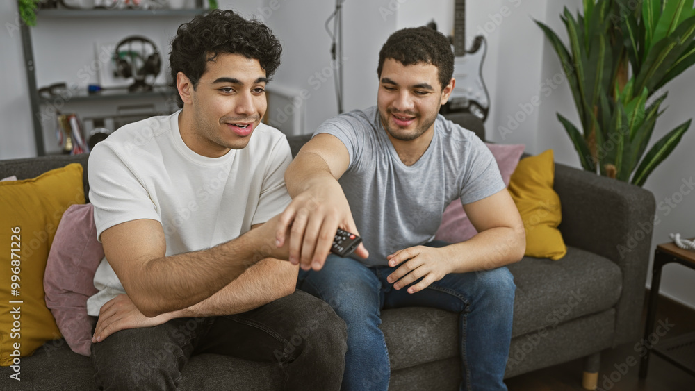 Two men sitting on a sofa indoors, casually dressed, watching tv with a ...
