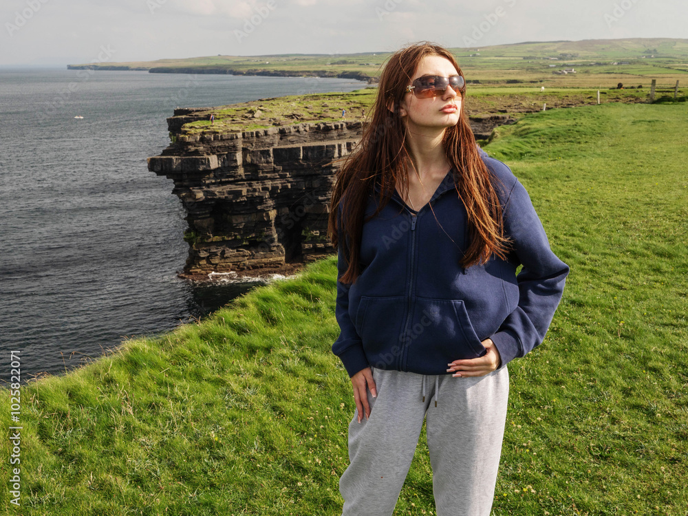 Beautiful teenager girl posing on a green field by a cliff, ocean and ...