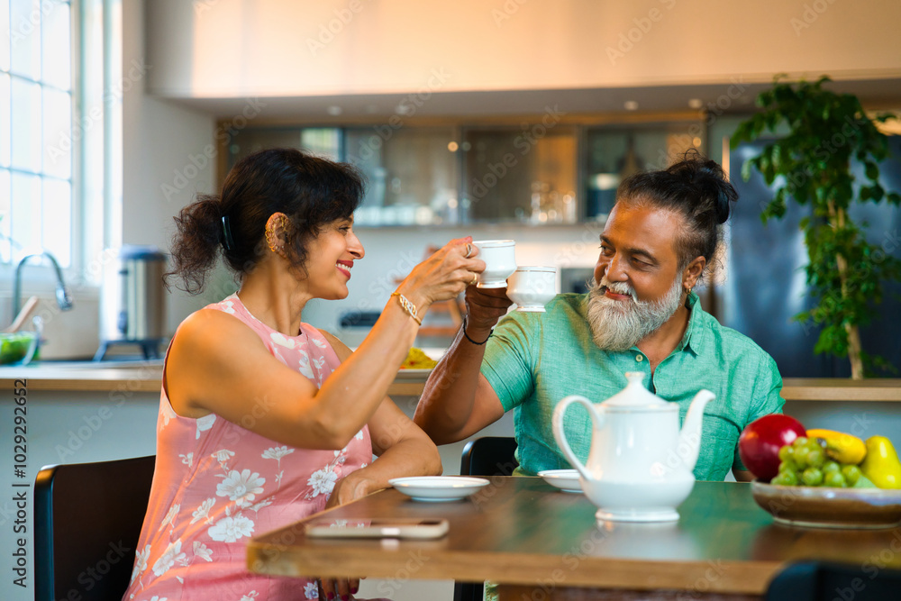 Indian Asian Middle aged couple toasting with tea cups at dining table ...