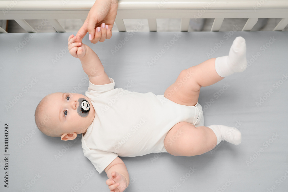 Baby with pacifier in mouth lies on back in crib and holds mom hand ...