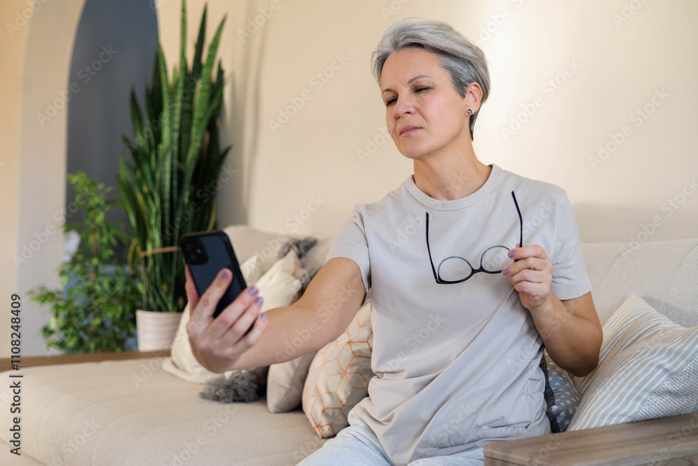 An elderly woman with gray hair and poor eyesight sits on the couch ...