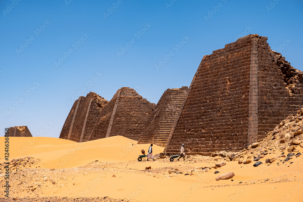Ancient pyramids of Meroe stand tall in the desert as two workers push ...