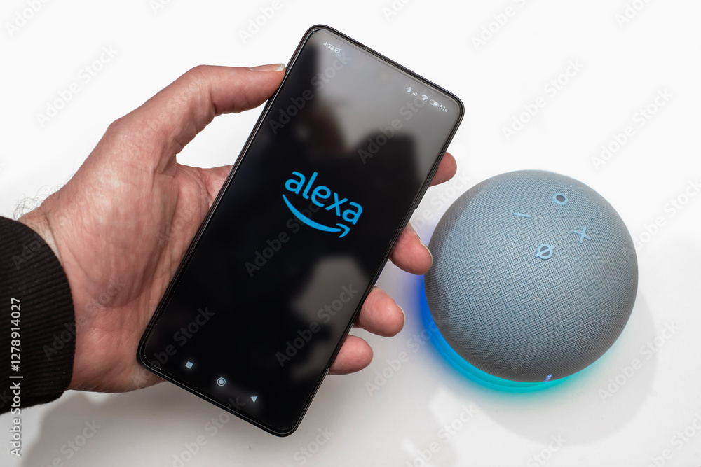 Mano sosteniendo un smartphone con el logo de Alexa junto a un altavoz ...