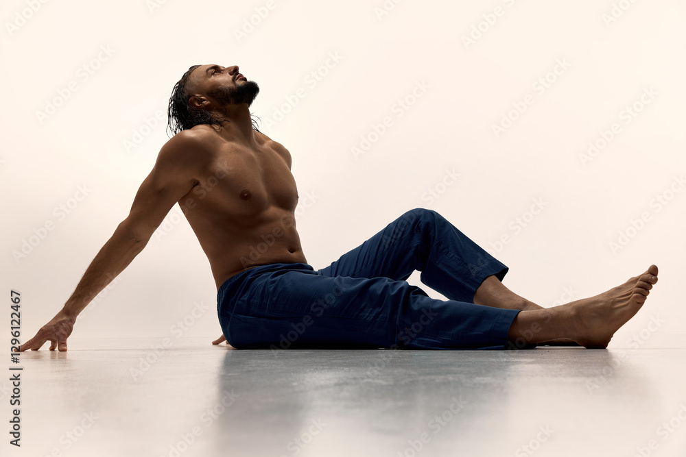 Young man with muscular upper body sitting shirtless on floor in loose ...
