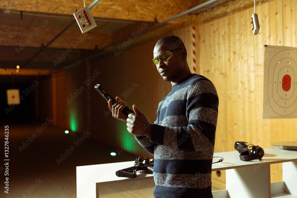 Man wearing protective goggles reloading gun with bullets in shooting range, following safety ...