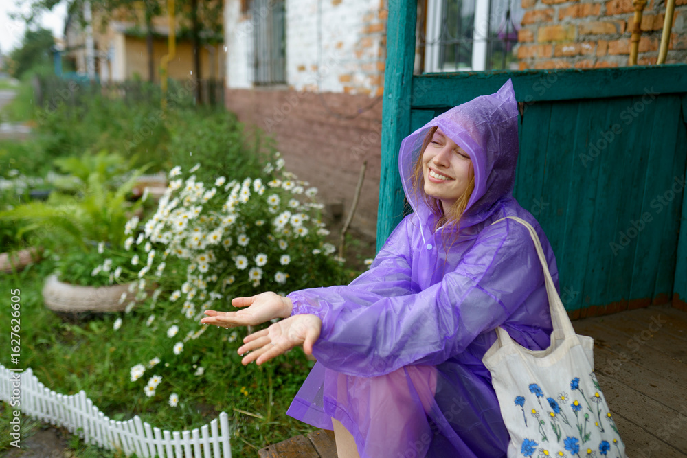 Smiling woman in purple raincoat enjoying rain outdoors near daisies ...