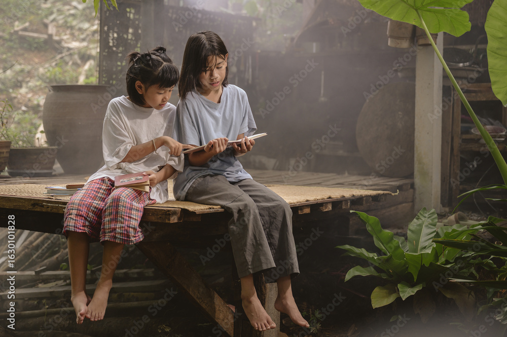 Two Asian children read together on a wooden porch in a rural Thai home ...