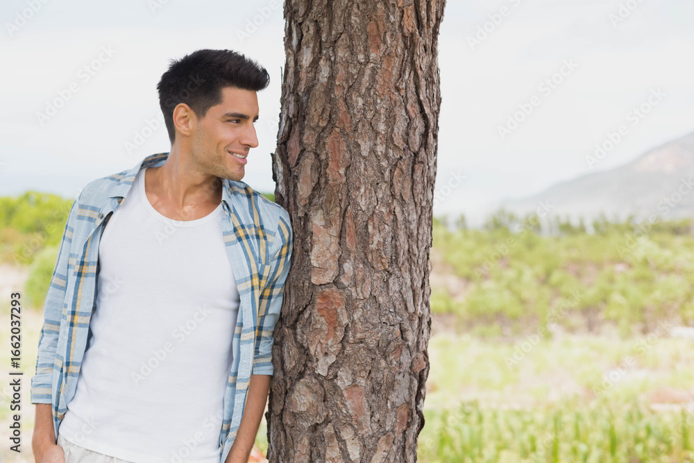 Man leaning against tree trunk in field amid low shrubs overlooking ...