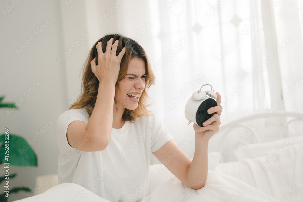 Frustrated woman sitting in bed, screaming at alarm clock after waking ...