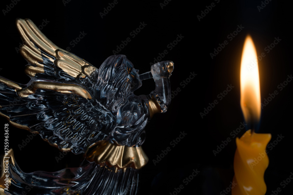 Glass angel with a trumpet beside a lit twist candle on black. Symbol ...