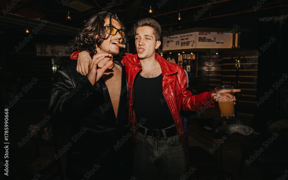 Two men share a lively moment in a dim bar, one in a red leather jacket ...