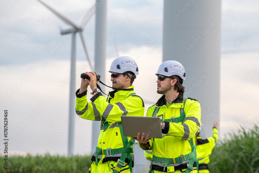 Two men in yellow jackets and hard hats are standing next to a wind ...