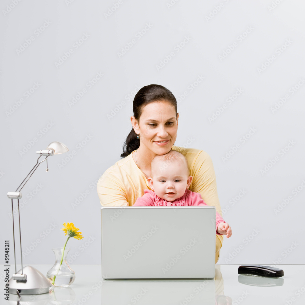 Working mother holding baby while typing on laptop at desk Stock Photo ...