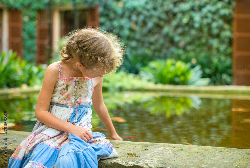 Little girl sitting by the pond with fish. Stock Photo | Adobe Stock