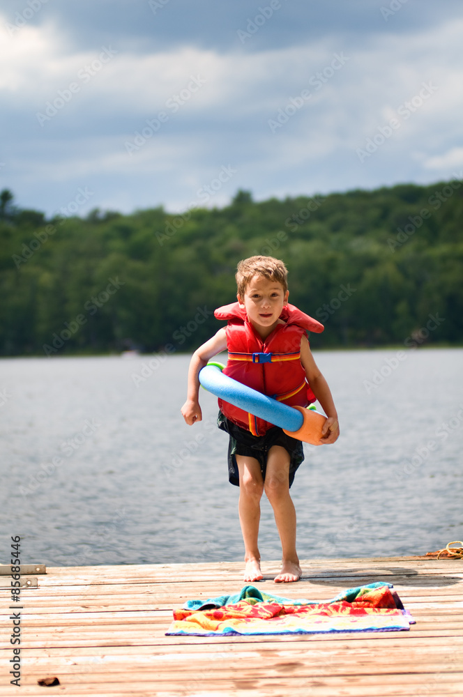 little boy standing on a dock by a lake wearing a life jacket and a ...