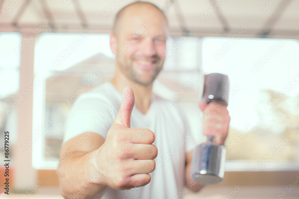 Fotka „Fitness man standing in a gym hold a dumbbell and giving thumb ...