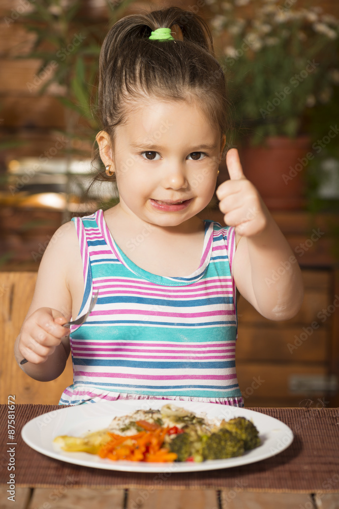 Little girl eating by itself broccoli, carrot and bell paper showing ok ...