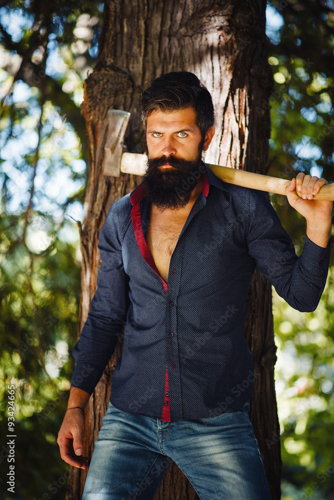 brutal bearded lumberjack with an ax at the tree in the forest lit by ...